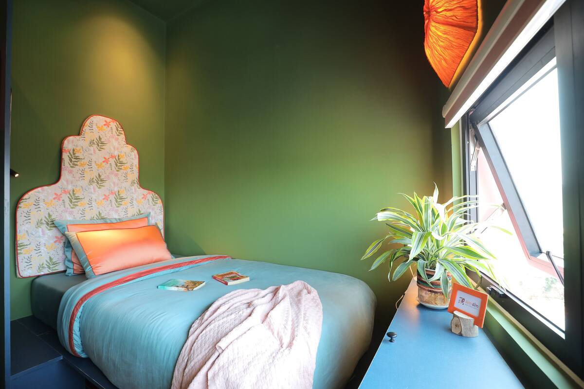 Uma cama com cabeceira florida, um cobre-leito azul e um cobertor rosa. Há uma planta em um parapeito de janela e uma pequena placa na janela.