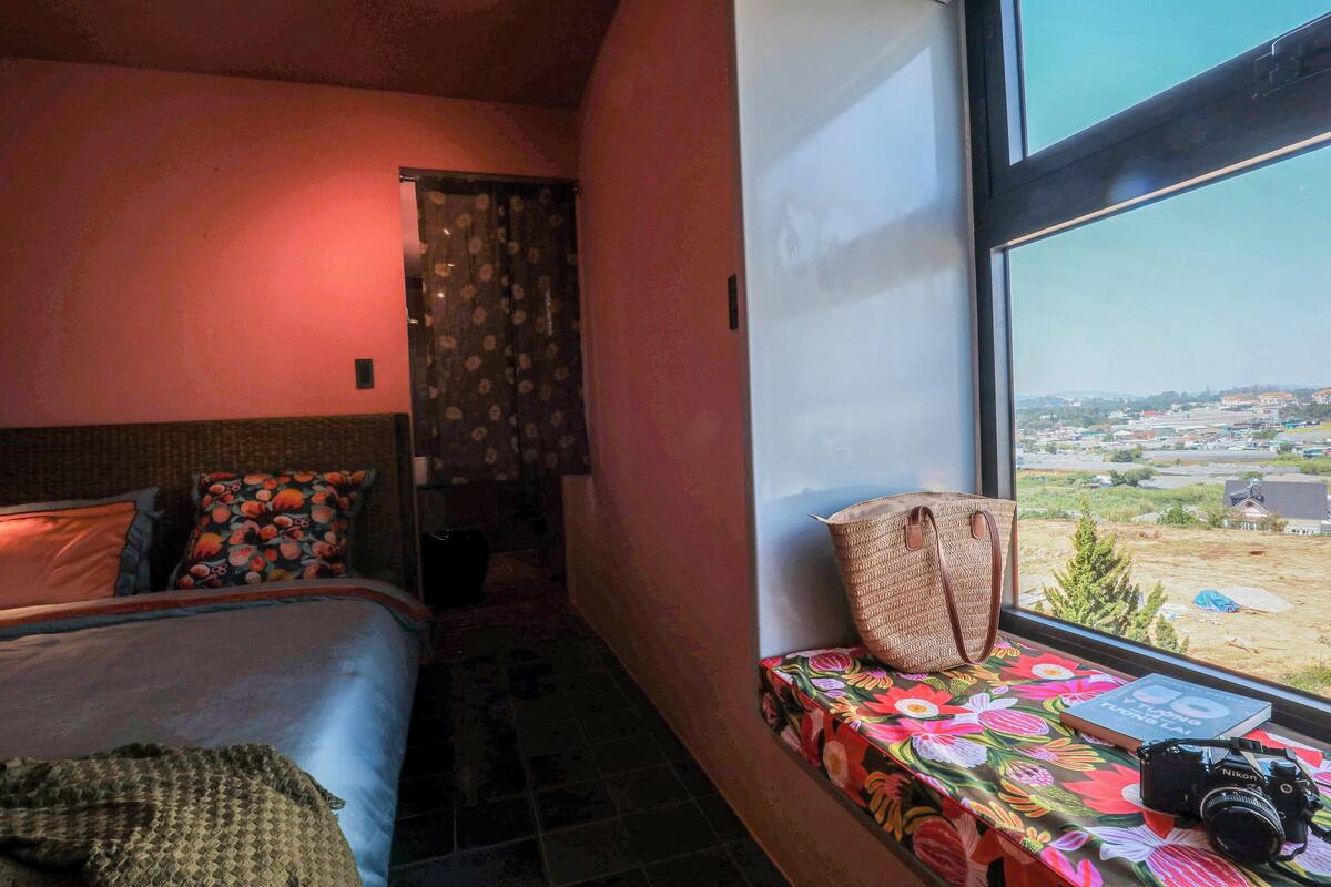 Quarto com cama, janela com vista para o campo, câmera e bolsa em um banco.