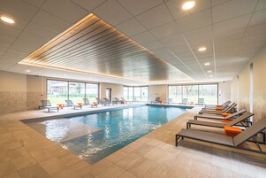 Uma piscina interior moderna com paredes de vidro transparente, espreguiçadeiras e chão em mosaico.