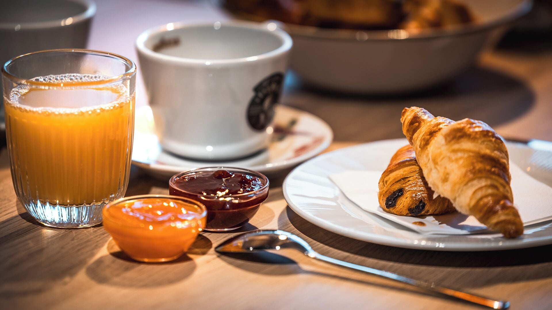 Uma mesa de madeira com café da manhã: um copo de suco de laranja, um croissant e duas pequenas tigelas de geleia.