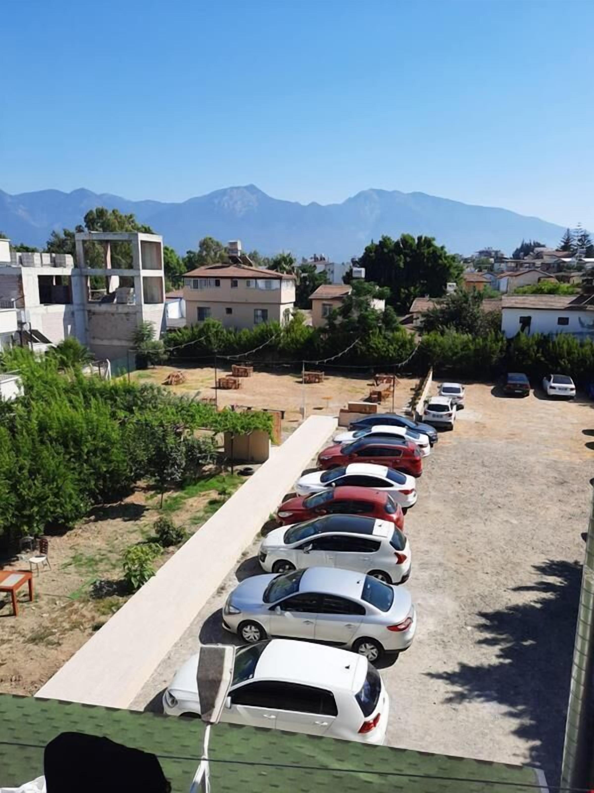Um parque de estacionamento com vários carros, um edifício com telhado plano e montanhas ao fundo.