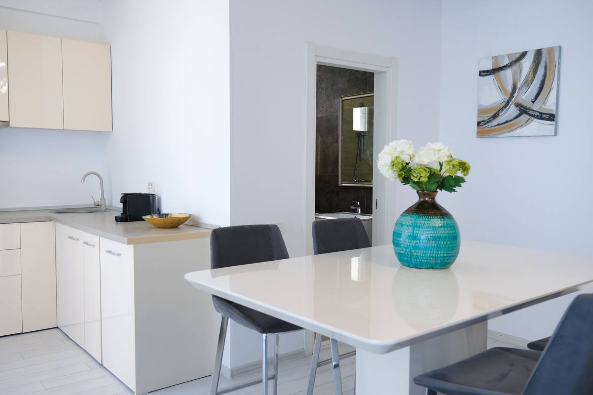 Cozinha moderna com armários brancos, bancada clara e pia. Há uma mesa de jantar com cadeiras e um vaso com flores.