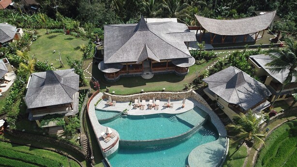 Vista aérea de um resort com piscina central, vários edifícios com telhados de palha e jardins bem cuidados.