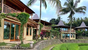 Um resort com várias construções de madeira, paredes de pedra e vegetação exuberante, incluindo palmeiras e um campo de arroz.
