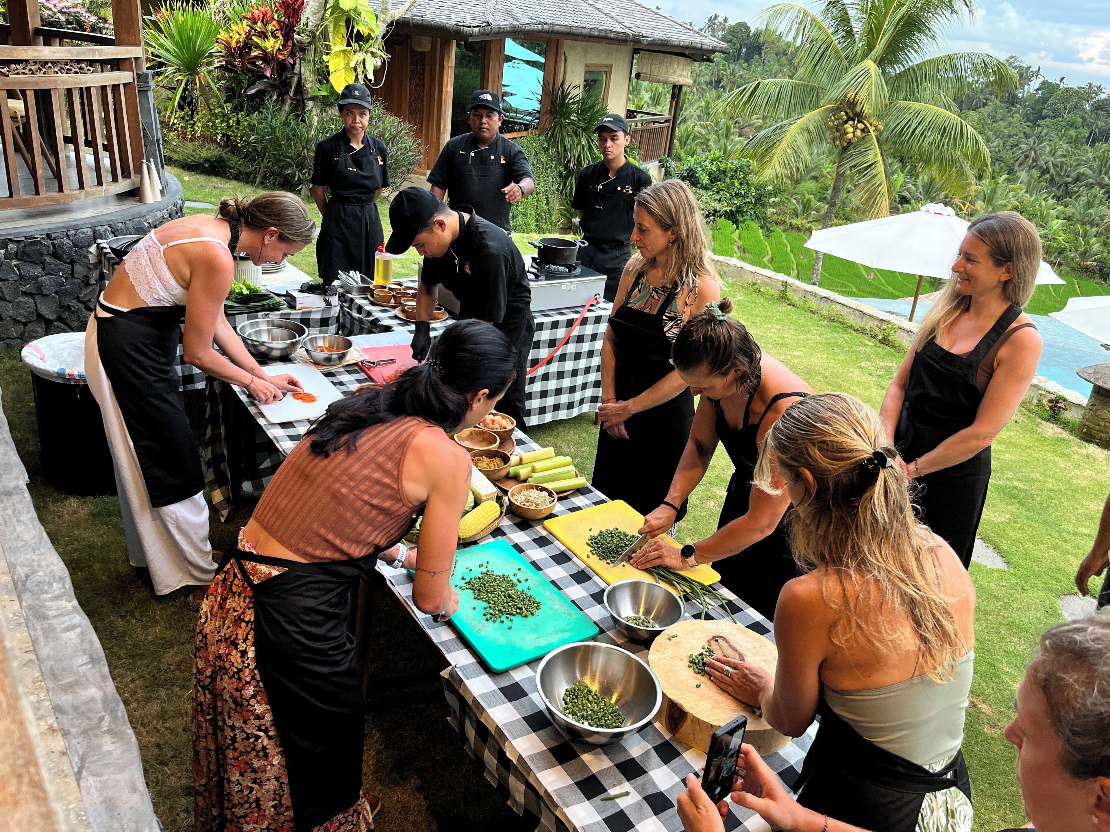 Um grupo de pessoas preparando comida ao ar livre, com toalha xadrez, piscina e vegetação exuberante ao fundo.