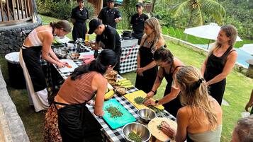 Um grupo de pessoas preparando comida ao ar livre, com toalha xadrez, piscina e vegetação exuberante ao fundo.