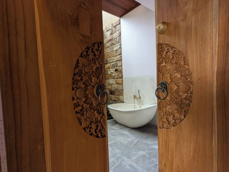 Uma porta de madeira com esculturas detalhadas, que dá acesso a um banheiro com banheira e parede de pedra.