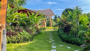 Um jardim tropical com um gazebo de palha e um caminho de pedra que leva a uma construção de madeira.