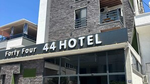 Um hotel moderno com fachada de pedra, um letreiro chamativo "44 HOTEL" e área externa com mesas e cadeiras amarelas.