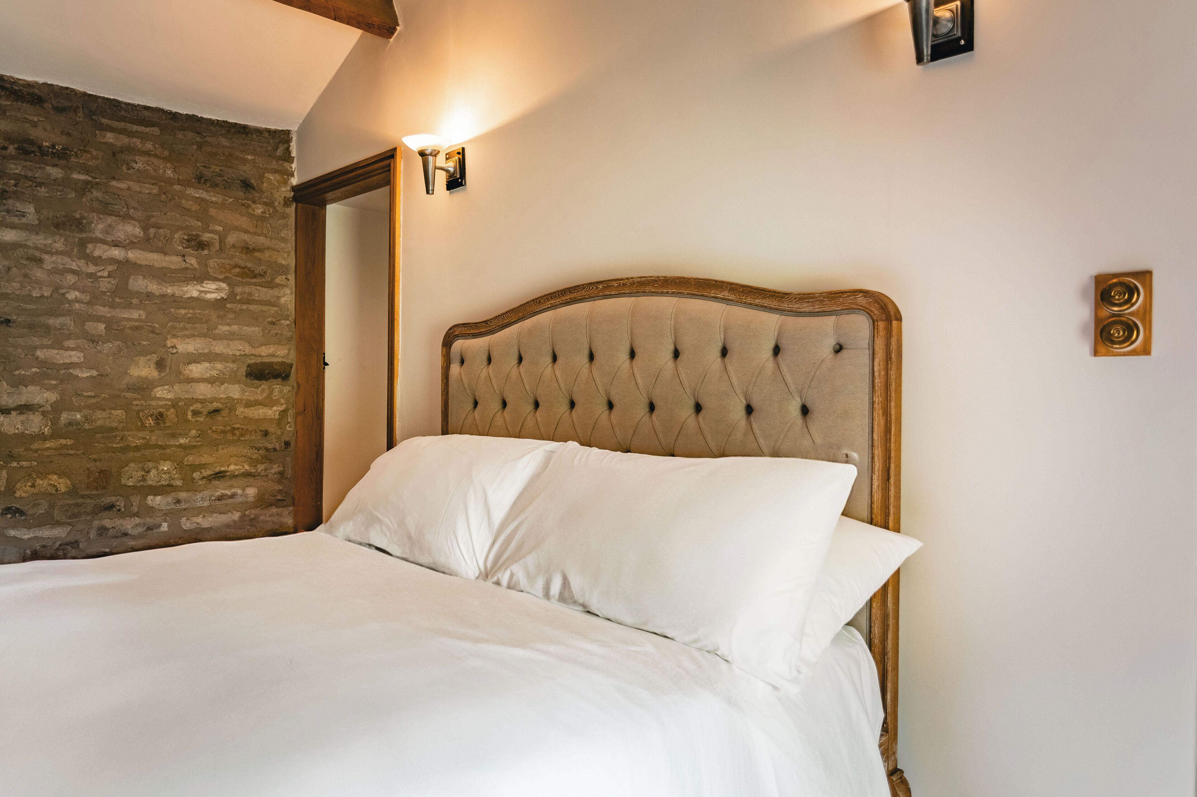 Cama com cabeceira estofada num quarto com paredes de pedra e teto de madeira.