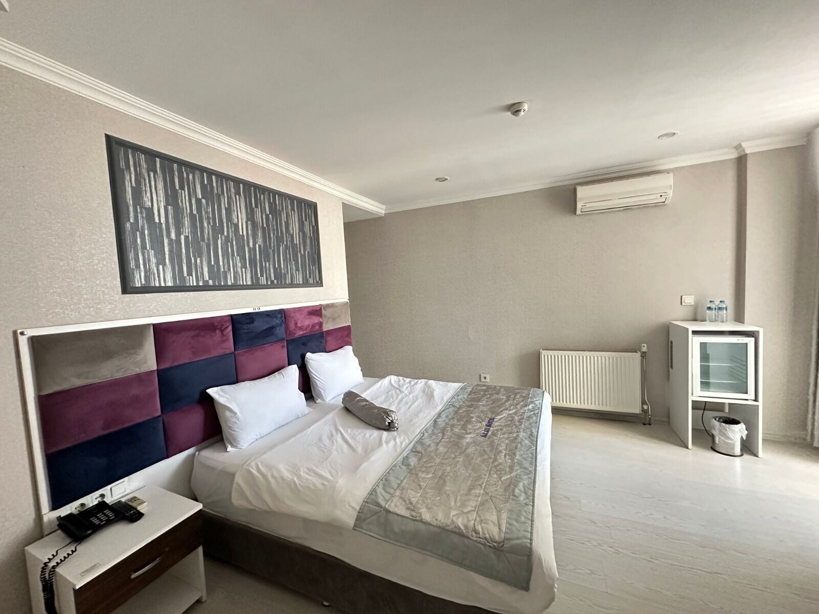 Quarto de hotel com uma cama grande, mesa de cabeceira com telefone e carregador, radiador e um móvel para televisão.