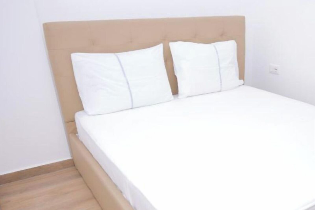 Uma cama com estrutura de madeira e roupa de cama branca num quarto com paredes e piso em tons claros.
