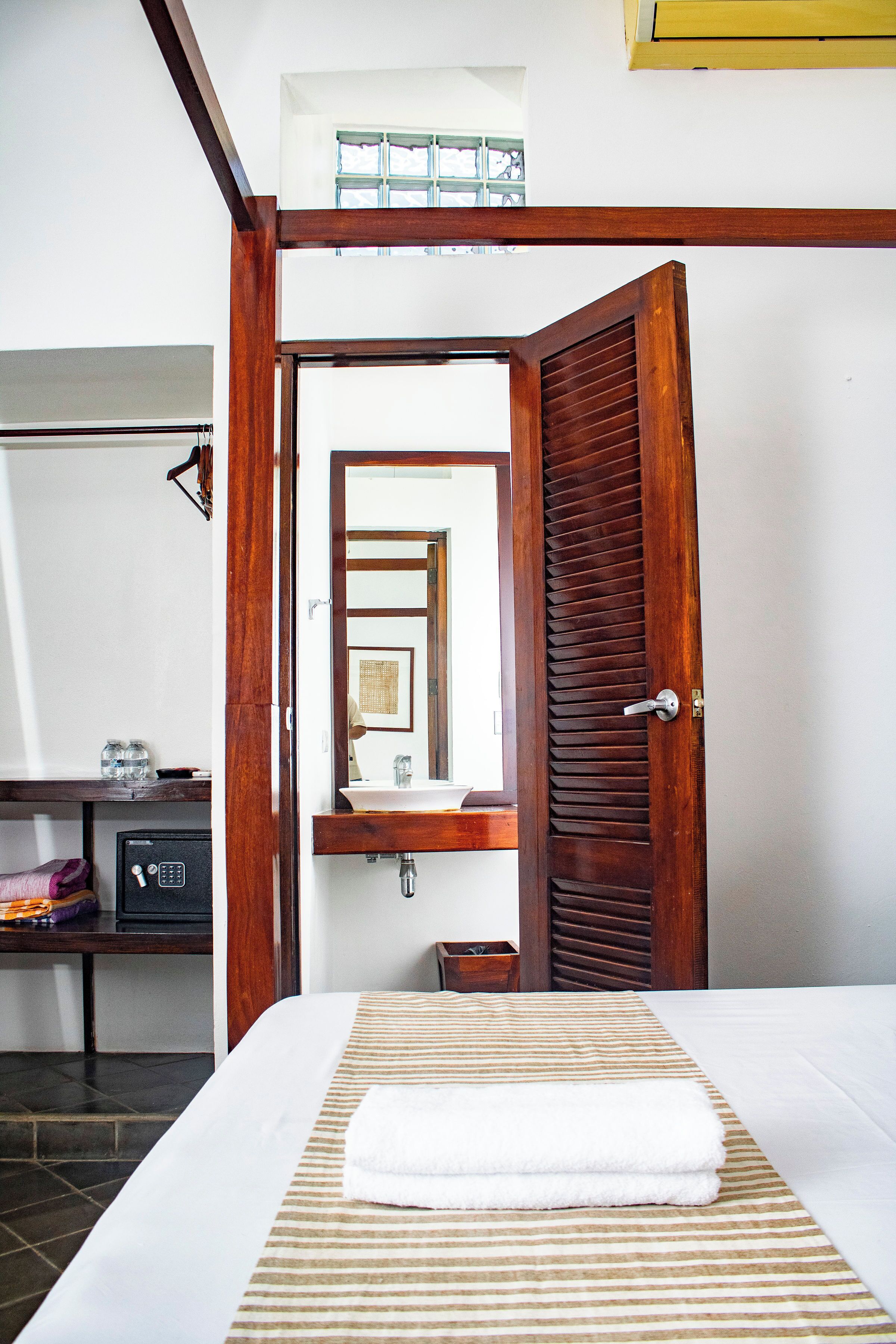 Quarto de hotel com cama, porta de madeira, espelho, mesa pequena e cofre.