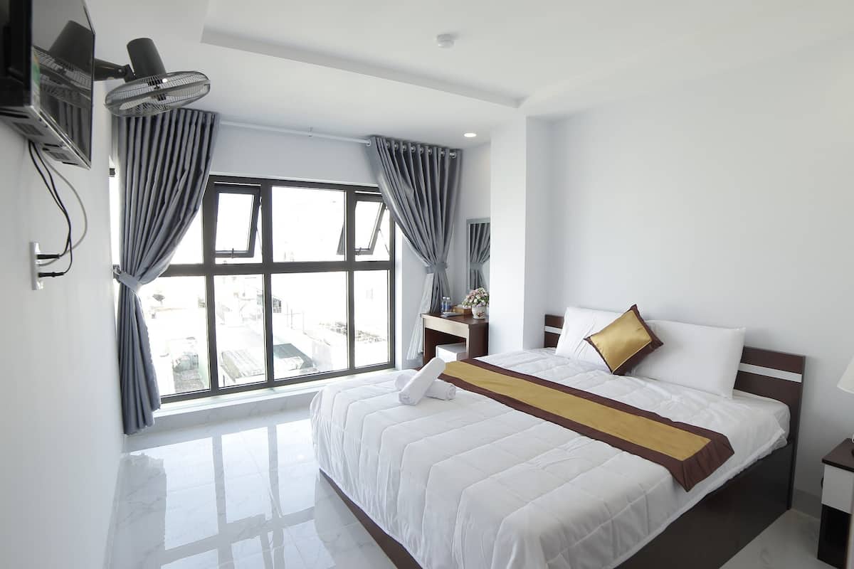 Quarto de hotel com uma cama grande, uma escrivaninha, uma cadeira, uma televisão fixada na parede e uma janela com cortinas.