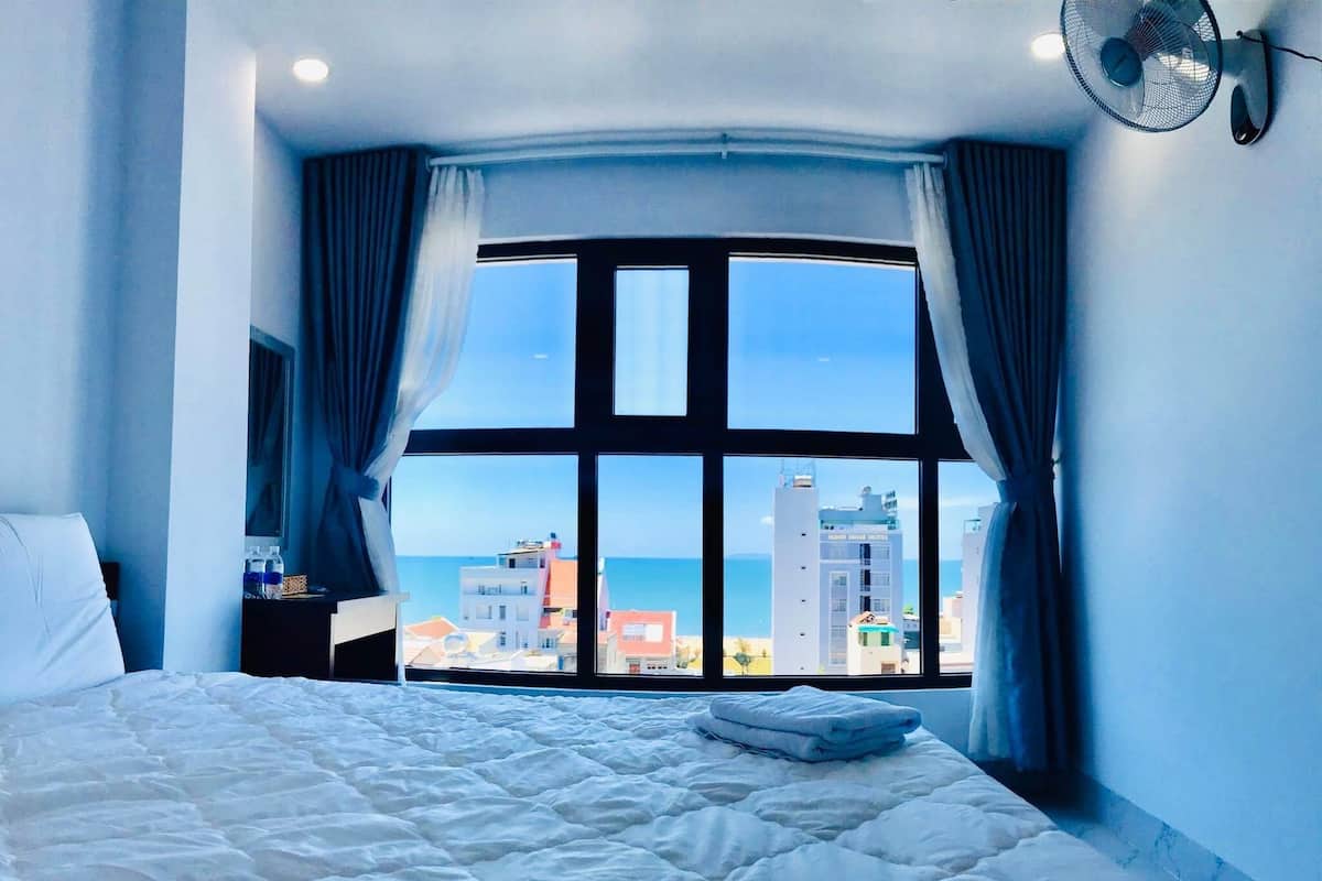 Quarto de hotel com uma cama grande, vista para a cidade e ventoinha no teto.