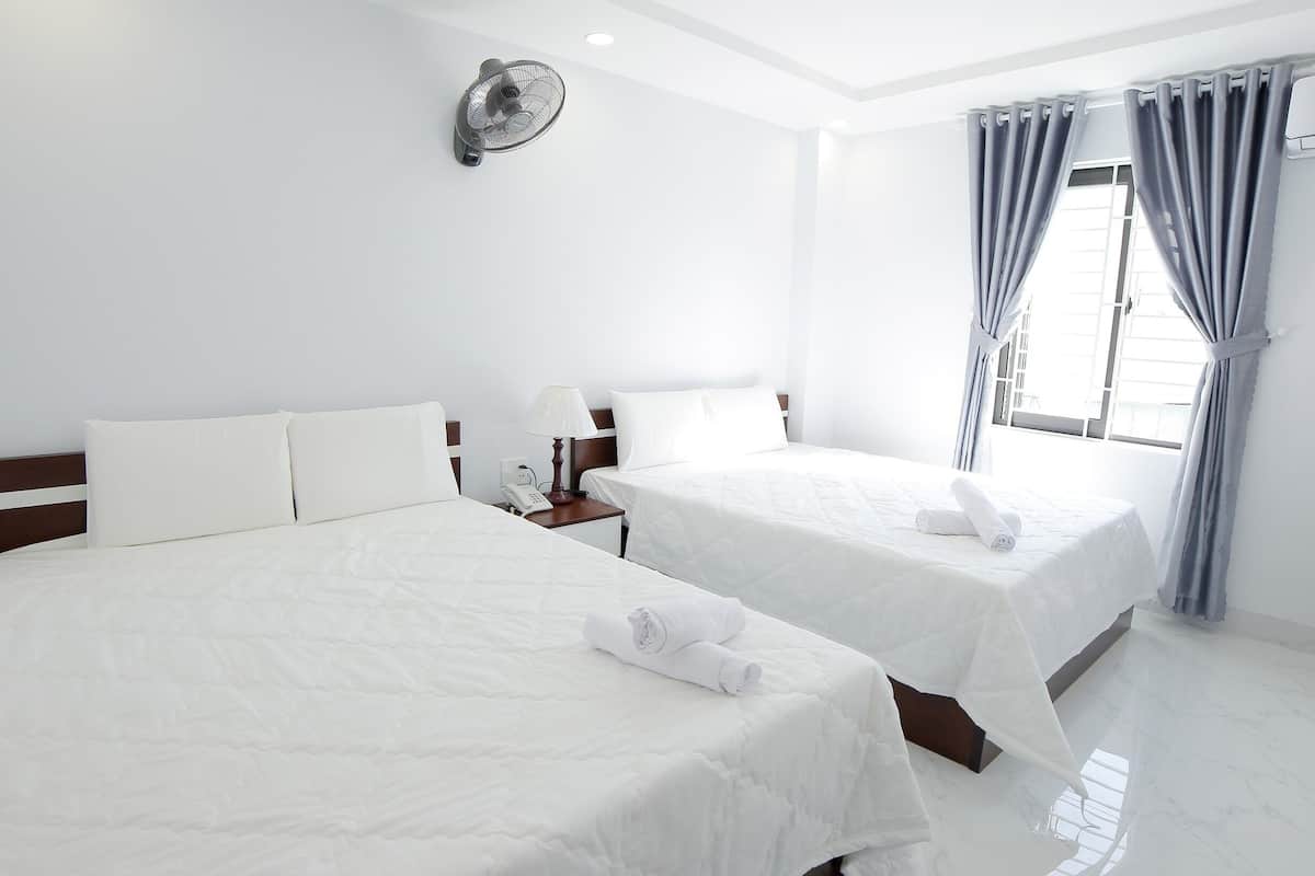 Quarto de hotel com duas camas, roupa de cama branca, mesa de cabeceira com abajur, ventoinha na parede e janela com cortinas.