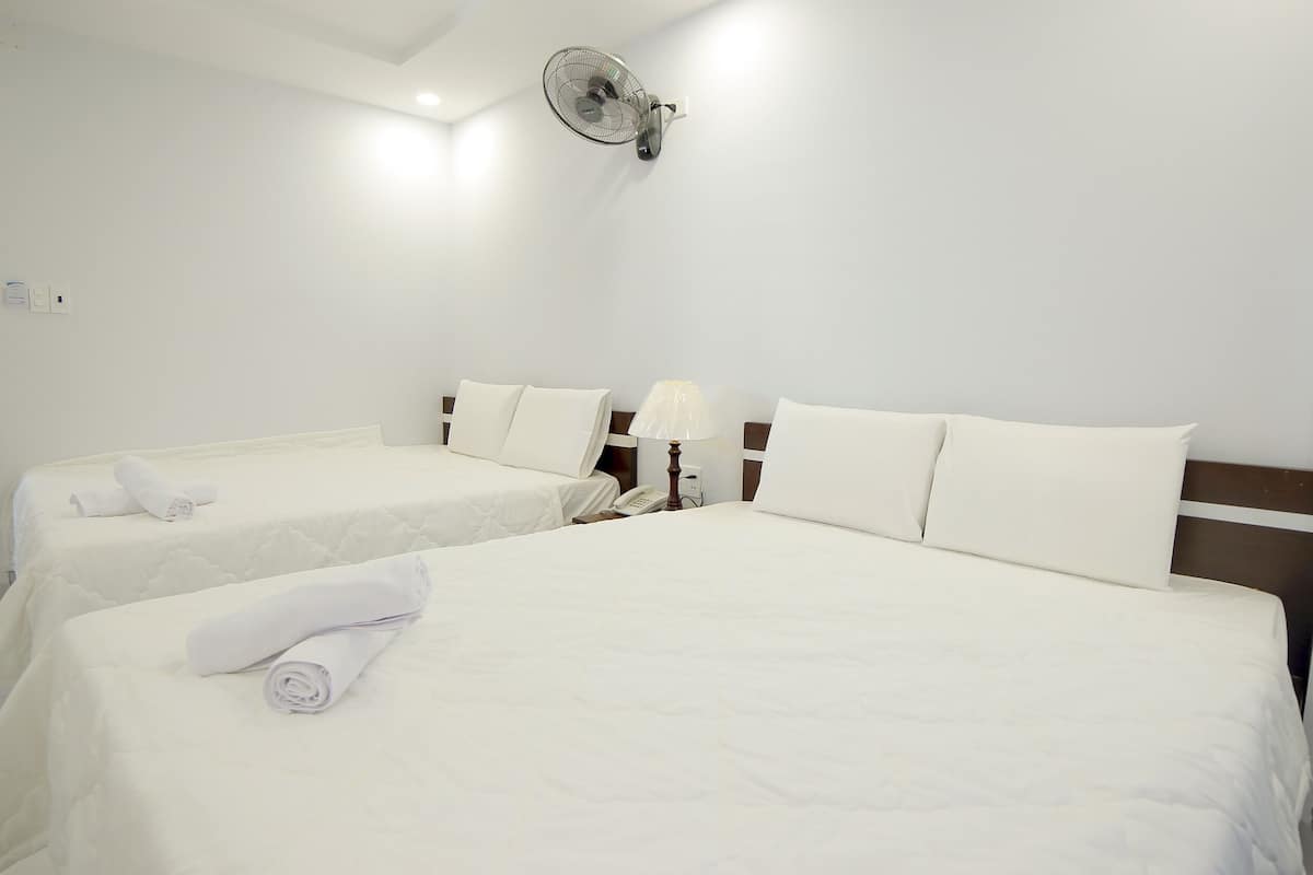 Quarto de hotel com duas camas, cada uma com roupa de cama e almofadas brancas, uma luminária de cabeceira e um ventilador fixado na parede.