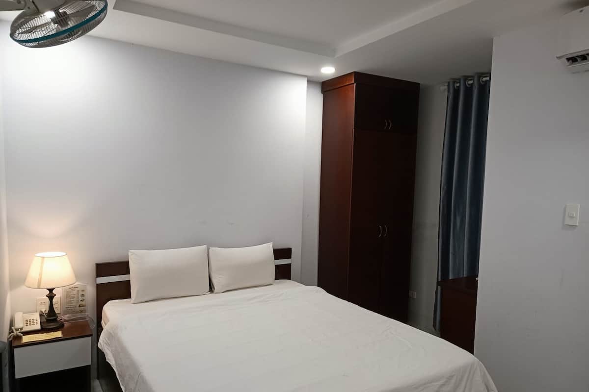 Quarto de hotel com cama, mesa de cabeceira com luminária, guarda-roupa e janela com cortinas.