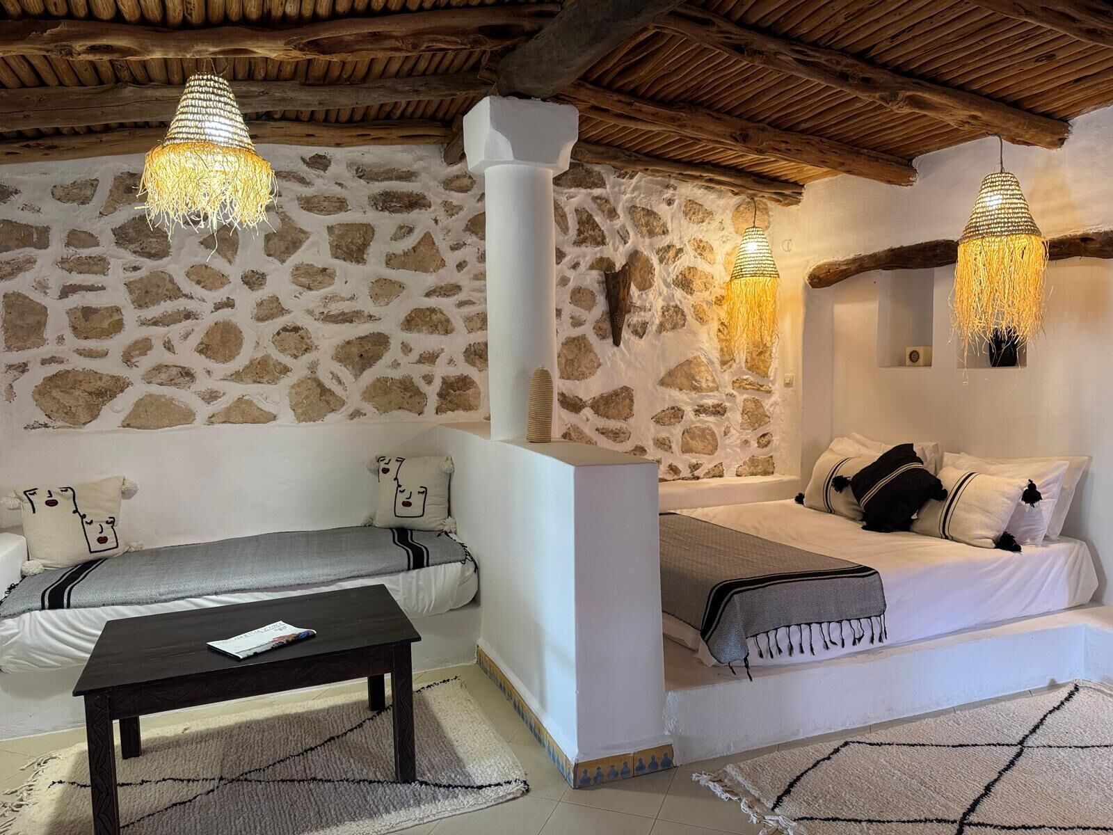 Um quarto acolhedor com parede de pedra, cama com manta listrada em branco e preto, sofá cinza com almofada, mesa de madeira e duas luminárias pendentes.