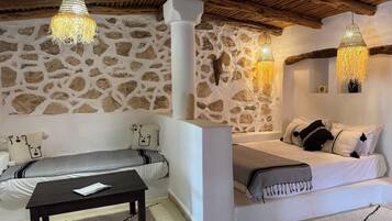 Um quarto acolhedor com parede de pedra, cama com manta listrada em branco e preto, sofá cinza com almofada, mesa de madeira e duas luminárias pendentes.