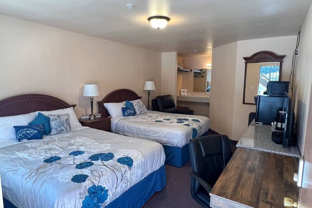 Quarto de hotel com duas camas, uma mesa de trabalho e uma cadeira. Há um espelho na parede, uma cafeteira e um cabideiro.