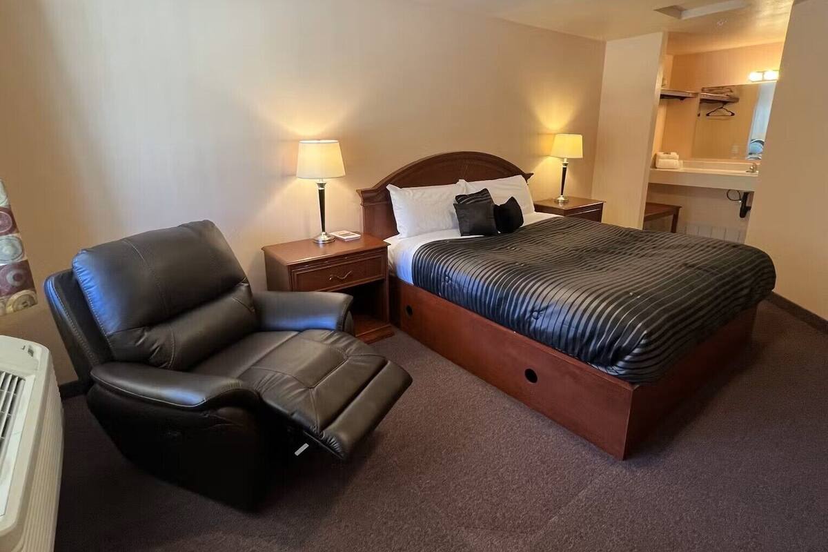 Quarto de hotel com uma cama, uma poltrona de couro, duas mesas de cabeceira com abajures, uma mesinha e um espelho.