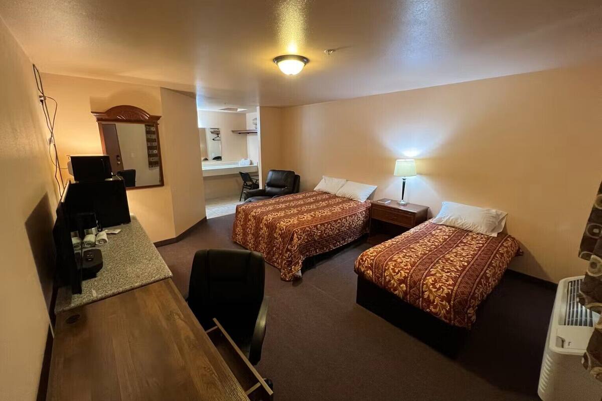Quarto de hotel com duas camas, uma escrivaninha, uma cadeira, uma luminária de mesa e iluminação no teto.