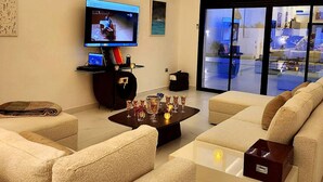 Uma sala de estar moderna com uma grande televisão de tela plana, sofás confortáveis, uma mesa de centro e vista através de amplas janelas.