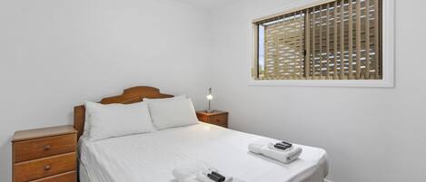 Uma cama bem arrumada com lençóis brancos, uma mesa de cabeceira de madeira com gavetas, um abajur e uma janela com persianas de madeira.