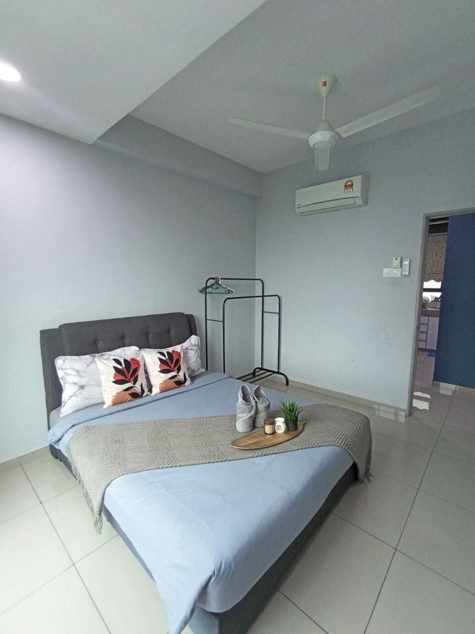 Quarto com cama, ventilador de teto, ar condicionado e uma pequena mesa com uma planta e uma xícara.