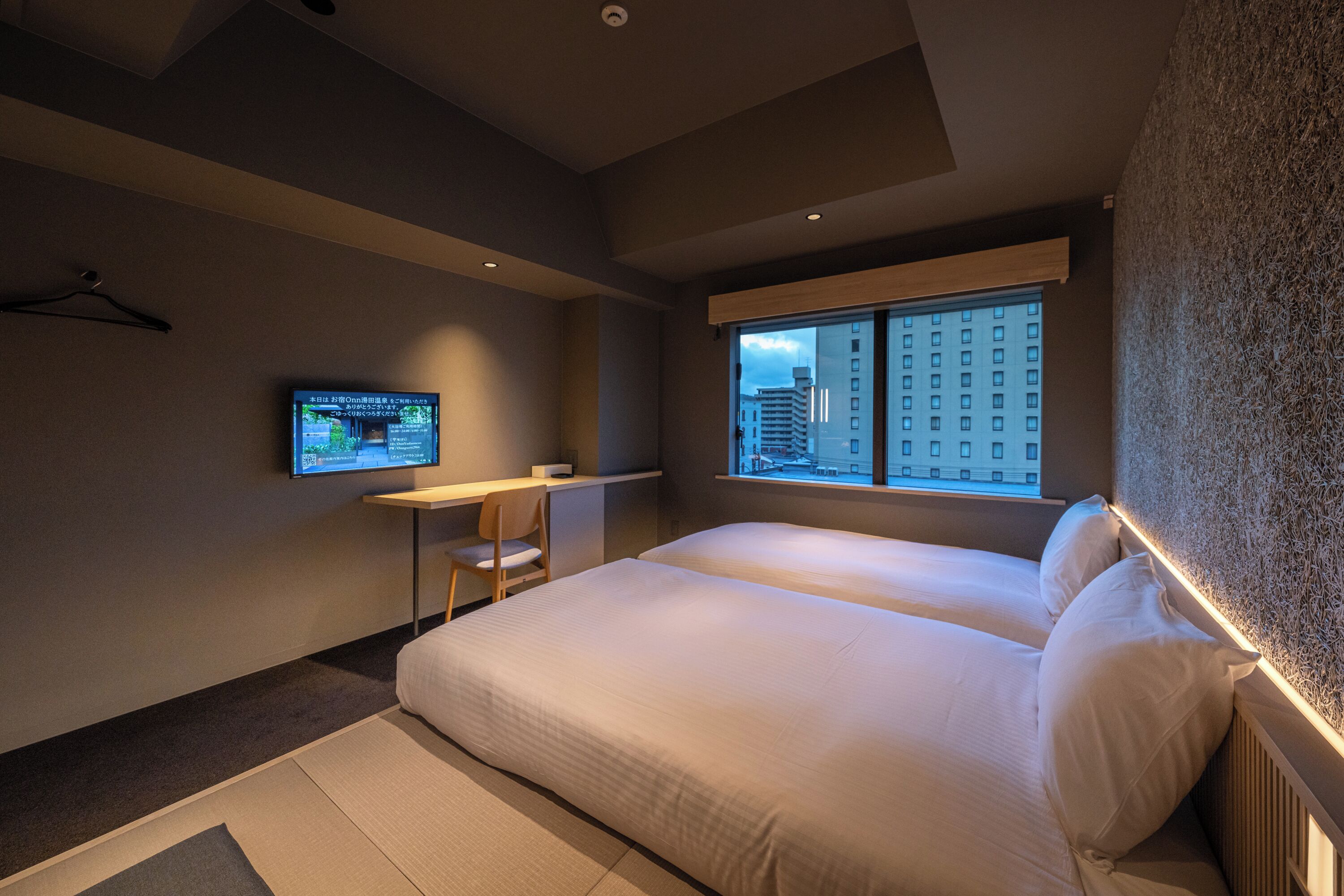 Quarto de hotel moderno com duas camas, uma escrivaninha e uma grande janela com vista para a cidade.