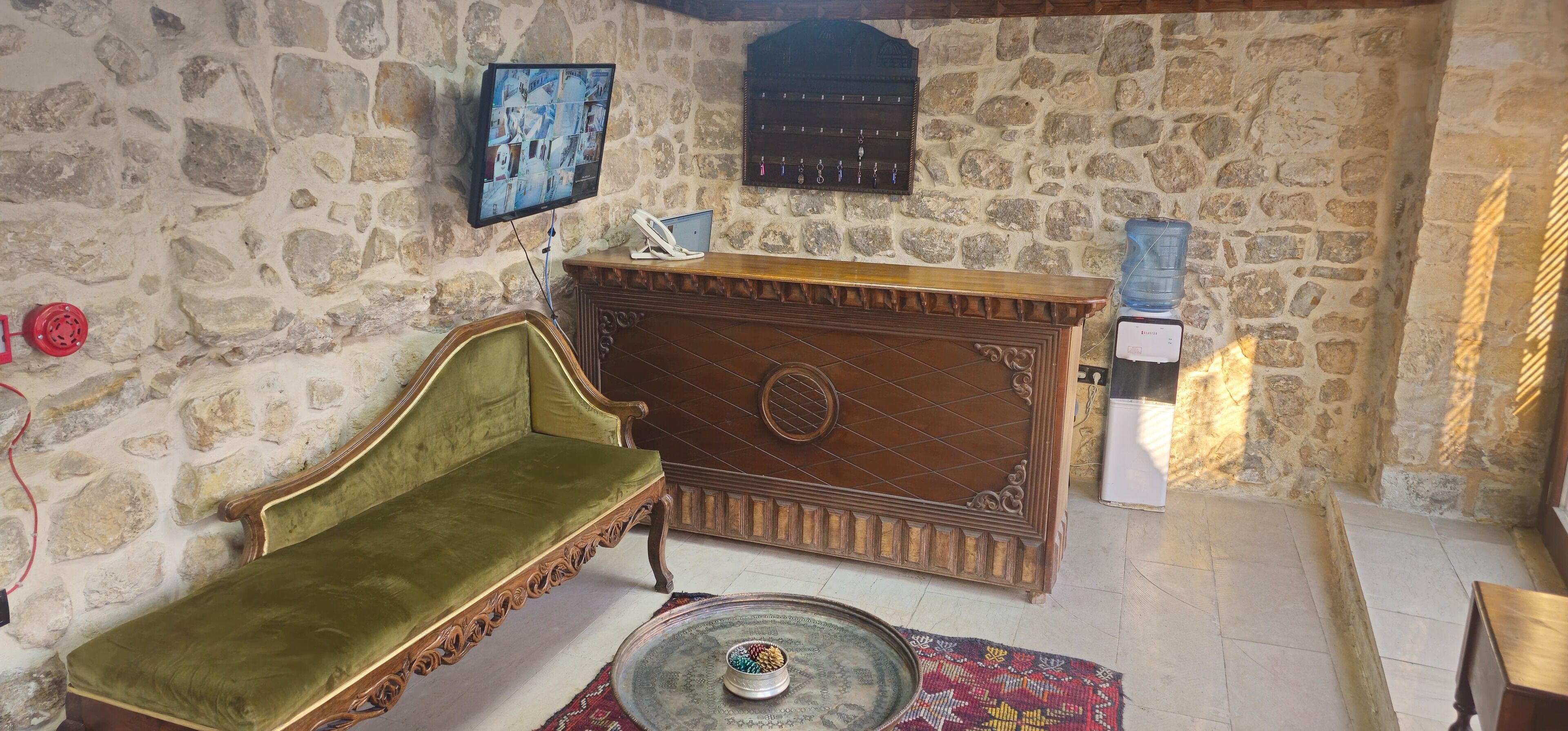 Quarto com paredes de pedra, um armário de madeira, um sofá verde, uma mesinha redonda com um tabuleiro decorativo e uma televisão na parede.
