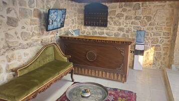 Quarto com paredes de pedra, um armário de madeira, um sofá verde, uma mesinha redonda com um tabuleiro decorativo e uma televisão na parede.