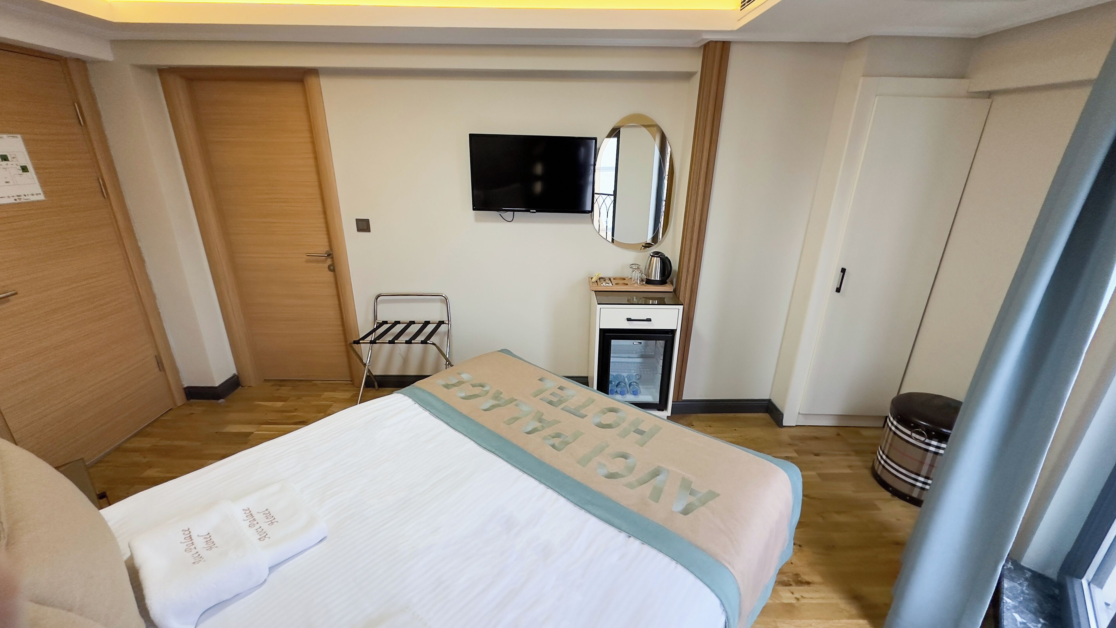 Quarto de hotel com cama, escrivaninha com refrigerador, televisão na parede e porta.