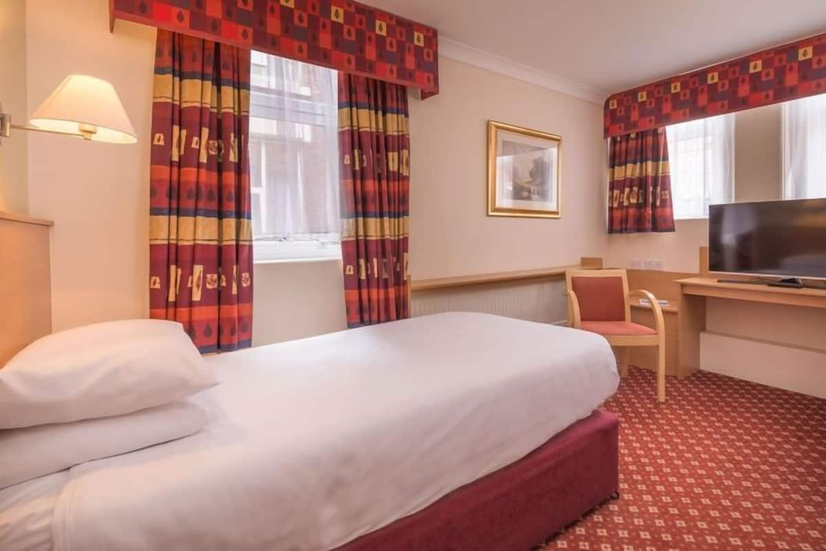 Quarto de hotel com cama, secretária e cadeira, televisão e cortinas.