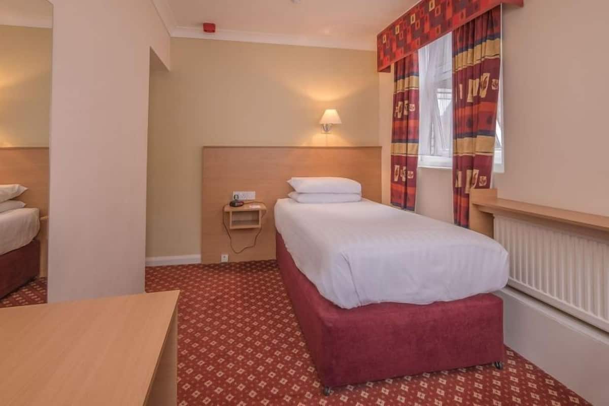Quarto de hotel com uma cama de solteiro, mesa de cabeceira, luminária e janela com cortinas estampadas.