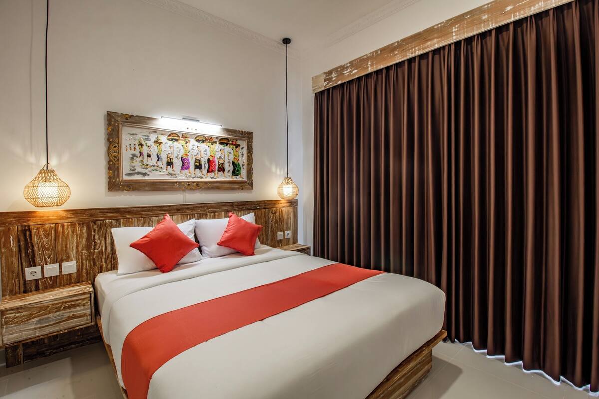 Quarto de hotel com uma cama grande, cabeceira de madeira, duas mesas de cabeceira, manta vermelha, um quadro na parede e uma janela com cortinas castanhas.