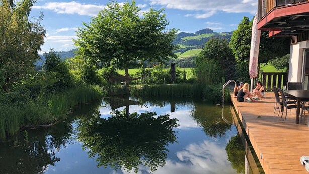 Área externa tranquila com um deck de madeira, um lago refletindo árvores e uma vista de colinas ondulantes ao fundo.