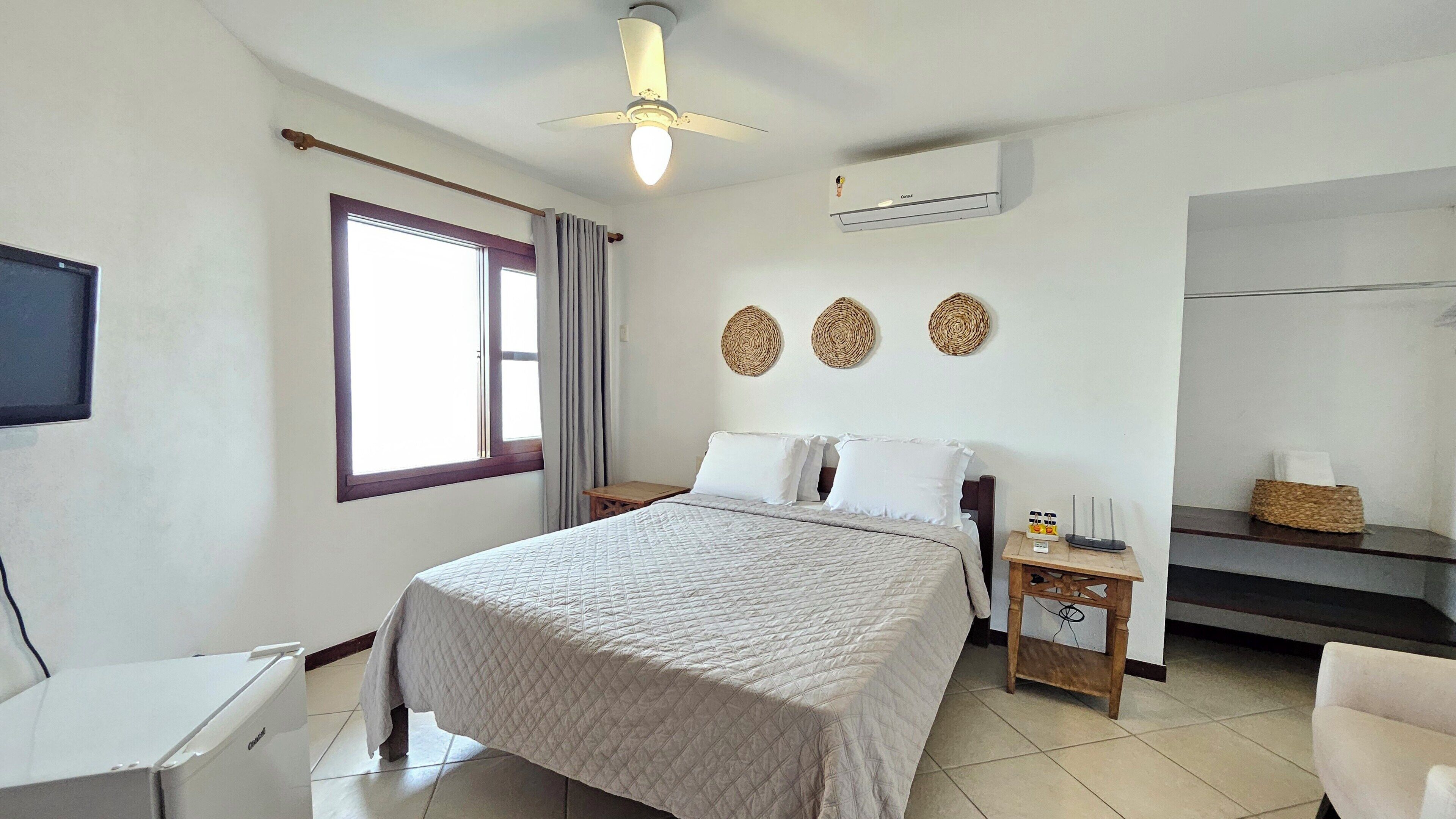 Quarto de hotel com cama, escrivaninha, cadeira, janela com cortinas, ventoinha no teto, ar condicionado e televisão de tela plana fixada na parede.