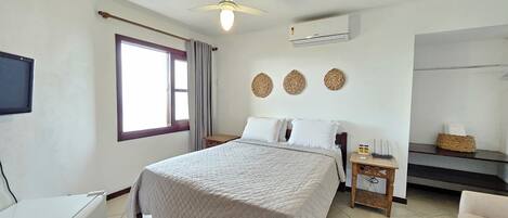 Quarto de hotel com cama, escrivaninha, cadeira, janela com cortinas, ventoinha no teto, ar condicionado e televisão de tela plana fixada na parede.