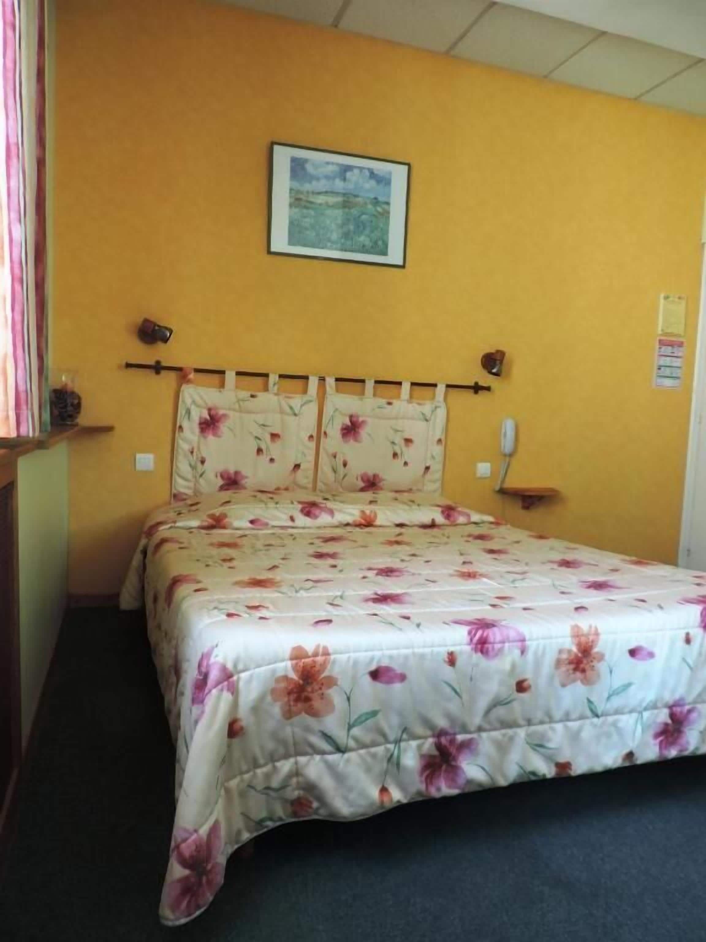 Uma cama de solteiro com colcha florida, um quadro pendurado na parede e um telefone na mesinha de cabeceira.
