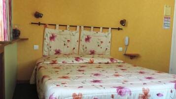 Uma cama de solteiro com colcha florida, um quadro pendurado na parede e um telefone na mesinha de cabeceira.