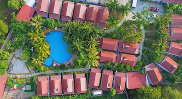 Vista aérea de um resort com uma piscina central rodeada por palmeiras e edifícios com telhados vermelhos.