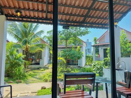 Um espaço ao ar livre coberto, com um banco e mesa, rodeado por vegetação exuberante e edifícios brancos com telhados vermelhos.