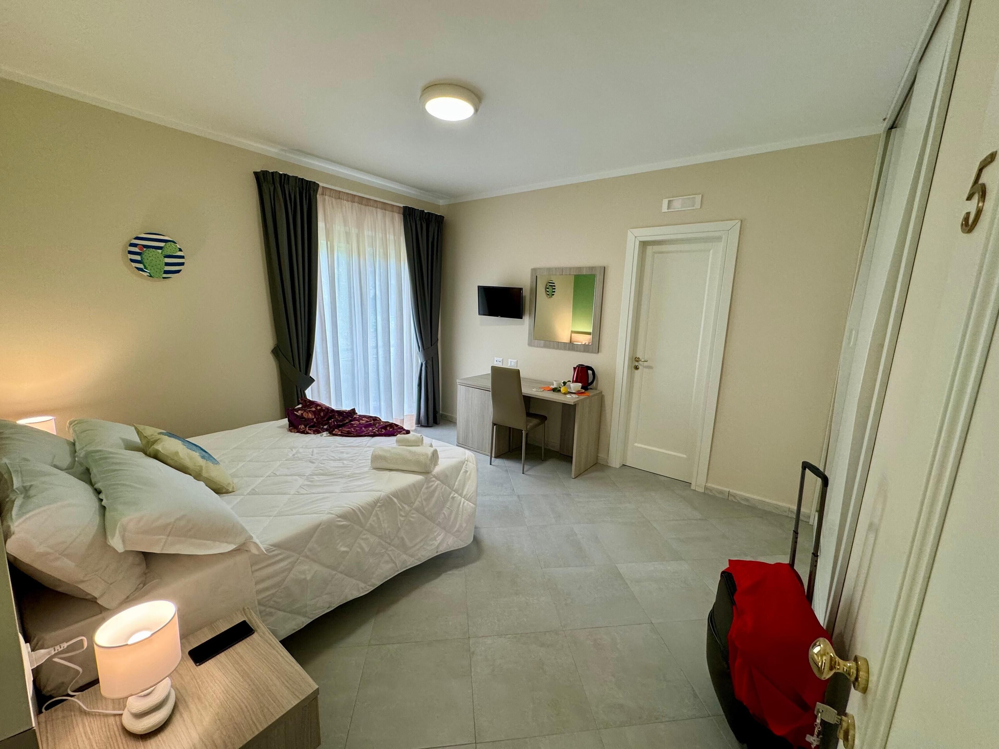 Quarto de hotel com cama, mesa de cabeceira com abajur, escrivaninha com cadeira, televisão fixada na parede e porta com placa de identificação.