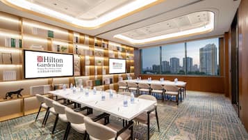 Uma sala de conferências do Hilton Garden Inn com uma tela grande exibindo o nome do hotel, vista para a cidade e um carpete com padrão geométrico.