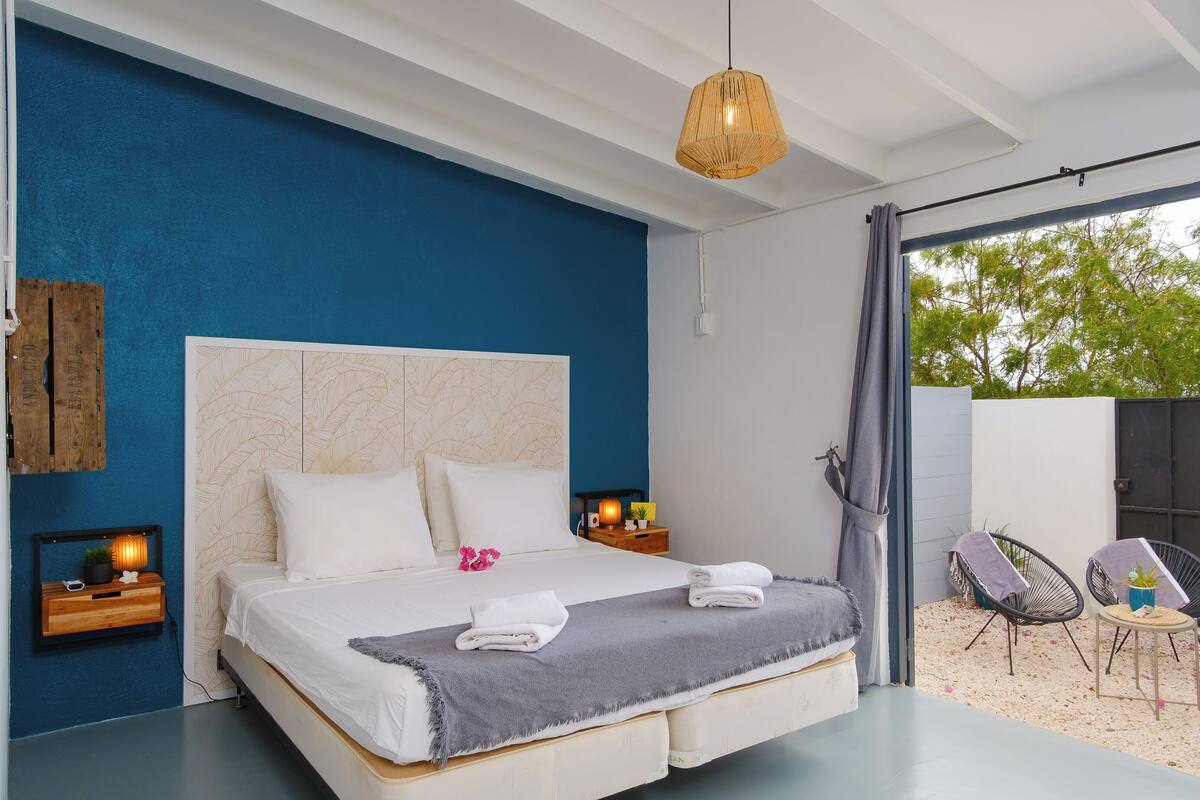 Quarto moderno com cama grande, cabeceira de madeira, mesas de cabeceira com abajures e vista para área externa com cadeiras e mesa através de uma porta deslizante.