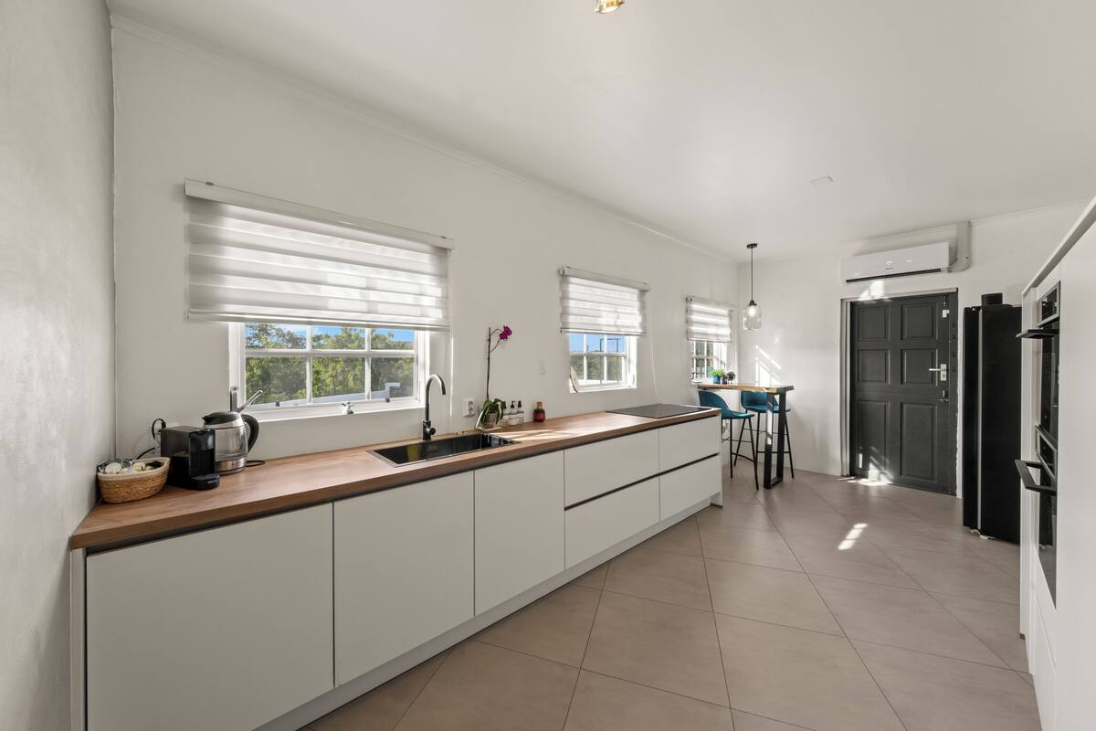 Cozinha moderna com armários brancos, bancada de madeira e pia embutida. Três janelas com persianas, um espaço de refeições com mesa e cadeiras, e uma porta preta.