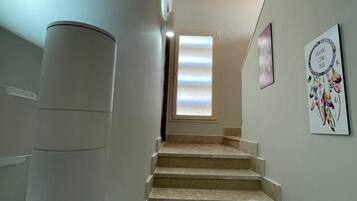 Uma escadaria com degraus bege conduzindo a um patamar, um objeto cilíndrico à esquerda e uma janela com persianas no topo.