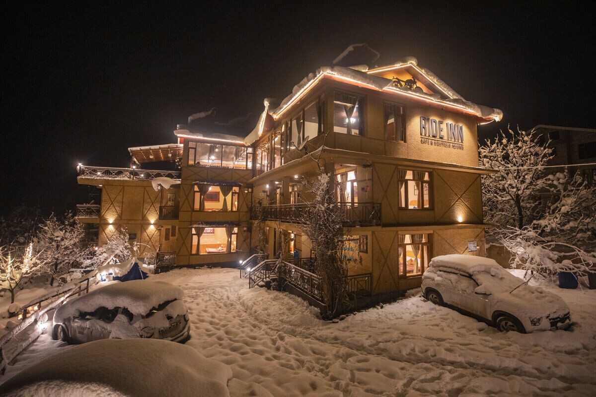Um hotel com vários andares, fachada acolhedora e janelas iluminadas, rodeado por neve e carros estacionados.